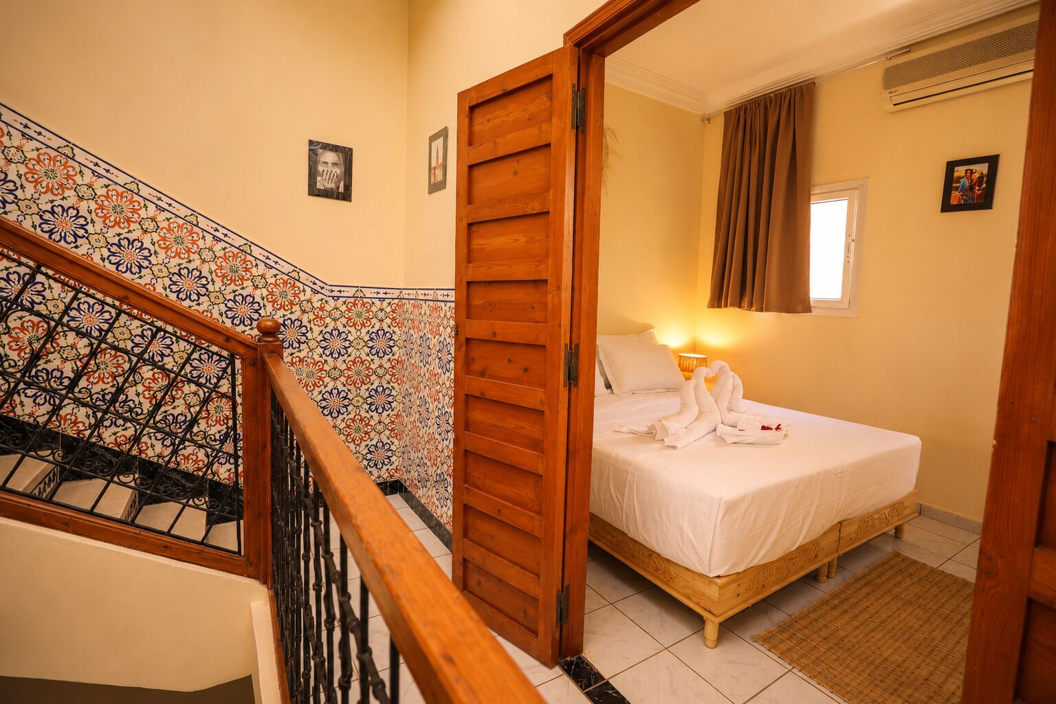 Quarto de hotel com uma cama, porta de madeira, escada com azulejos estampados e uma pequena janela com cortinas.