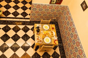 Uma mesa de jantar com pratos, copos e uma cesta de palha, sobre um chão quadriculado preto e branco, com uma parede estampada ao fundo.