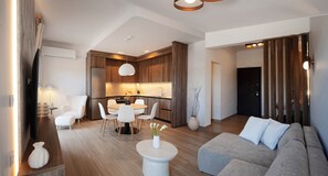 Uma sala de estar moderna com um sofá cinzento, uma área de jantar com mesa e cadeiras brancas e uma cozinha com armários de madeira e forno embutido.