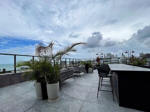 Terraço no telhado com área de estar, plantas em vasos e vista para o horizonte urbano e o oceano.