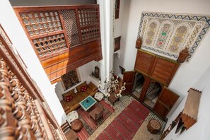 Interior tradicional com teto alto, separação em madeira treliçada, tapeçaria estampada e um lustre central.
