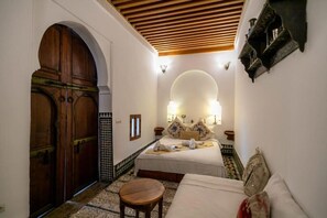 Quarto com porta de madeira grande, cama com colcha branca, mesa redonda pequena e prateleira fixada na parede.