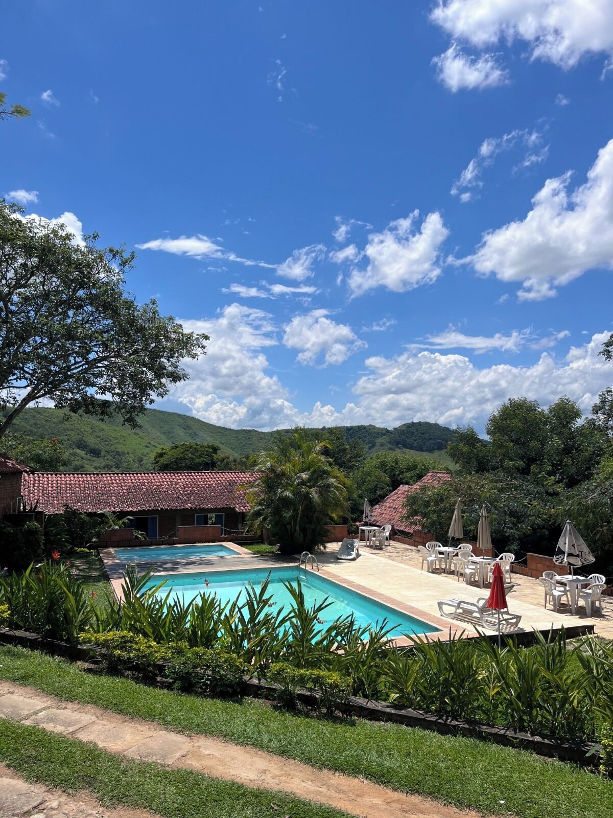 Uma área de piscina com céu claro, rodeada por vegetação e um edifício com telhado de telhas vermelhas.
