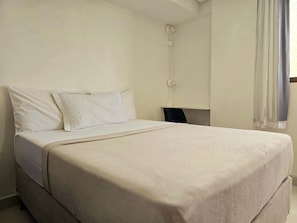 Uma cama com colcha e almofadas brancas, uma mesa de cabeceira com abajur e uma janela com cortinas.