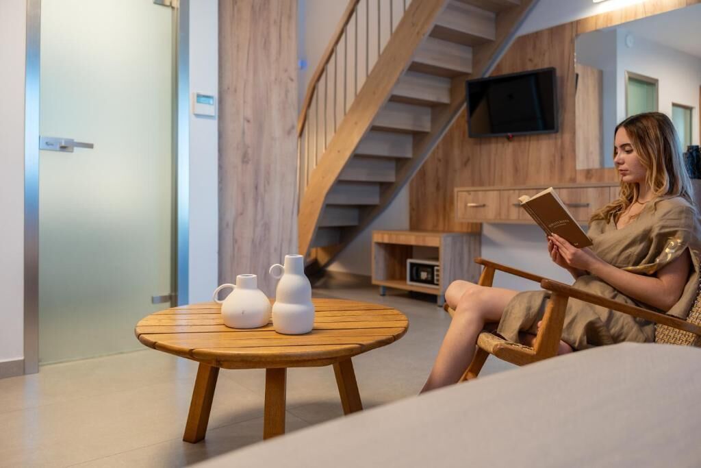 Um interior moderno com uma escada de madeira, uma mesa redonda de madeira com vasos brancos e uma pessoa lendo um livro.