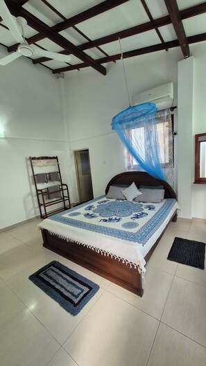 Quarto com cama, ar condicionado pendurado, ventoinha no teto, cabideiro de madeira e janela com cortina.