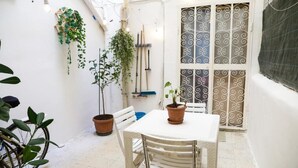 Um pequeno pátio com mesa e cadeiras, plantas em vasos e uma janela com grades decorativas de metal.