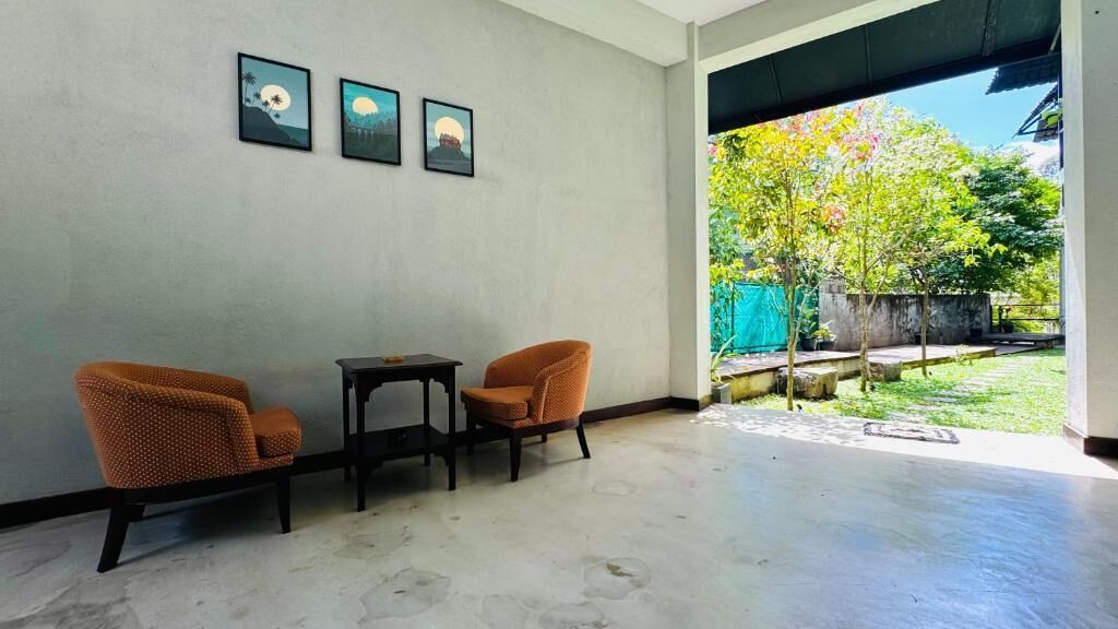 Quarto com duas cadeiras e uma mesa pequena, piso de concreto e vista para um jardim com árvores e uma cerca.