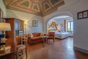Quarto de hotel com cama, sofá, mesa e cadeira. Teto com padrões decorativos e janela com cortinas.