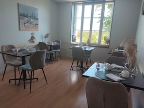 Uma sala de jantar com várias mesas e cadeiras, uma grande janela com vista para o verde e um quadro na parede.