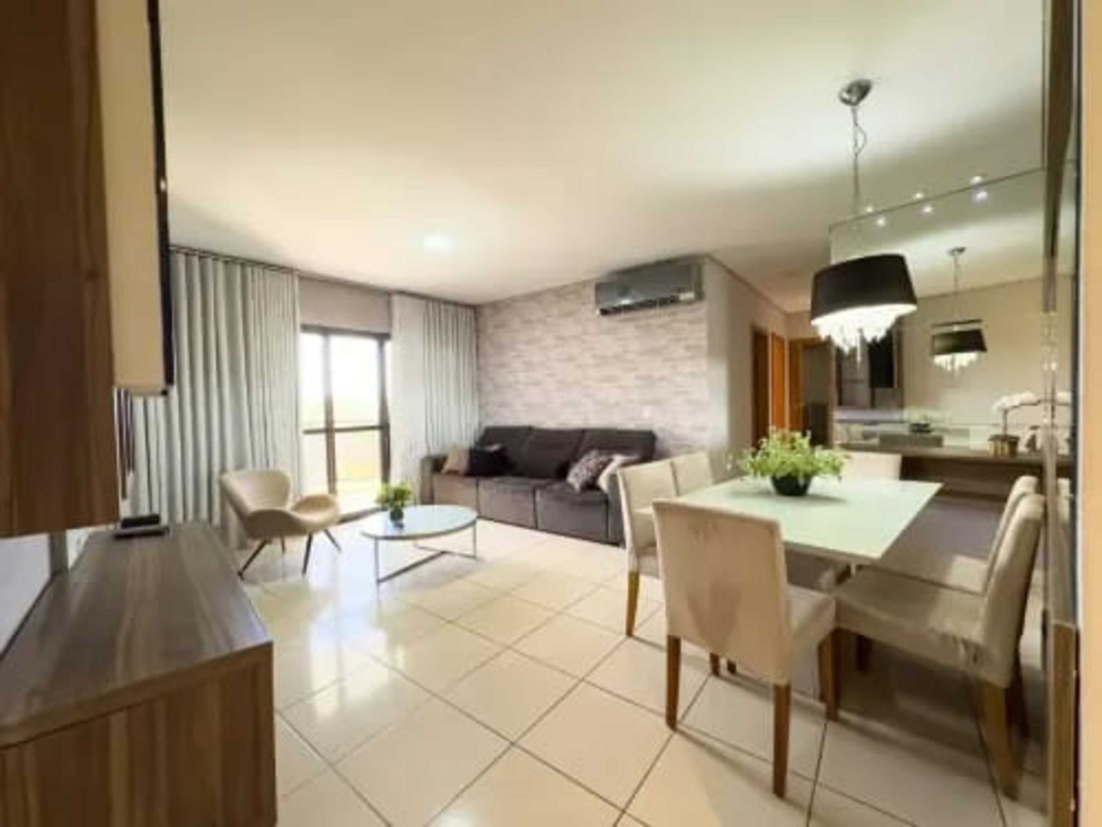 Uma sala de estar moderna com sofá, mesa de jantar com cadeiras e área de cozinha com luminária suspensa.