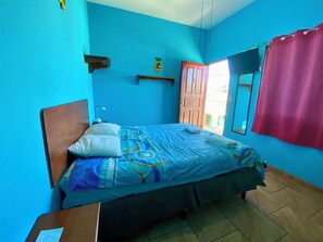 Quarto com cama, cabeceira de madeira, mesa de cabeceira, prateleira na parede, porta, janela com vista e cortina vermelha.