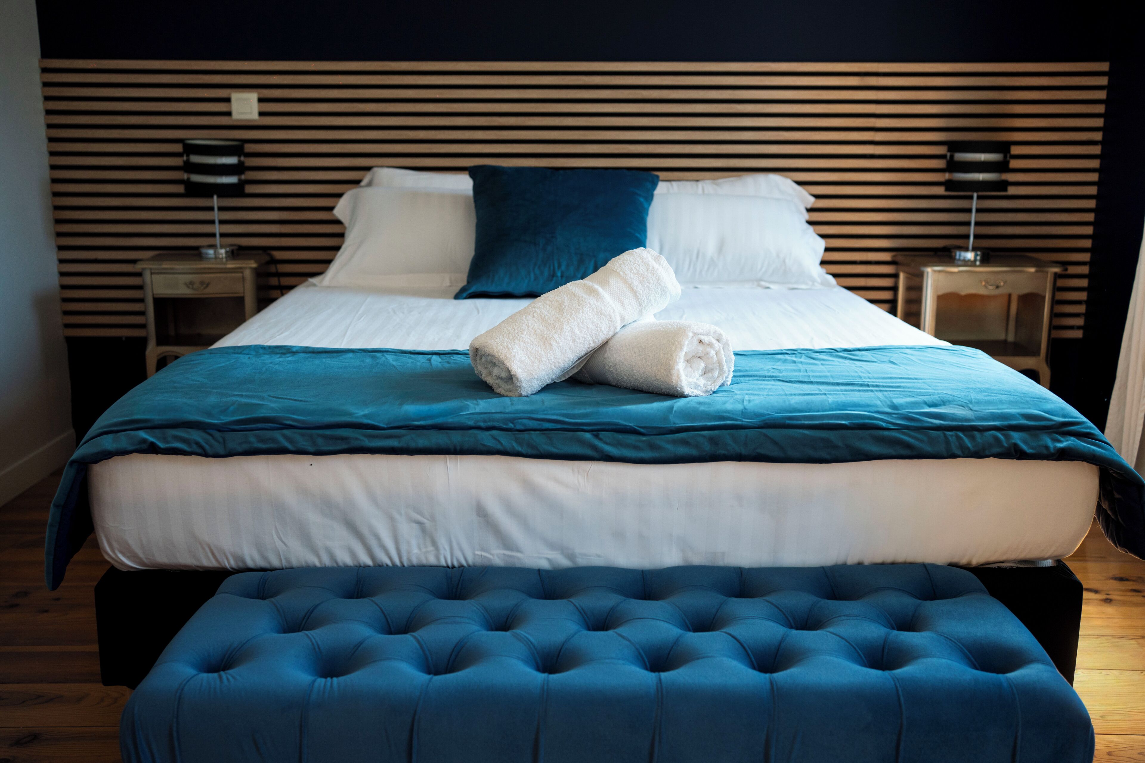 Cama bem arrumada com cabeceira azul, roupa de cama branca e azul, e duas toalhas enroladas sobre a cama.