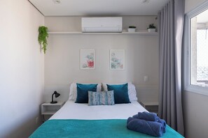 Quarto moderno com cama, almofadas, abajur, janela com cortinas, ar condicionado e duas obras de arte na parede.