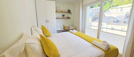 Quarto de hotel com cama, escrivaninha, cadeira e janela com vista para edifícios.