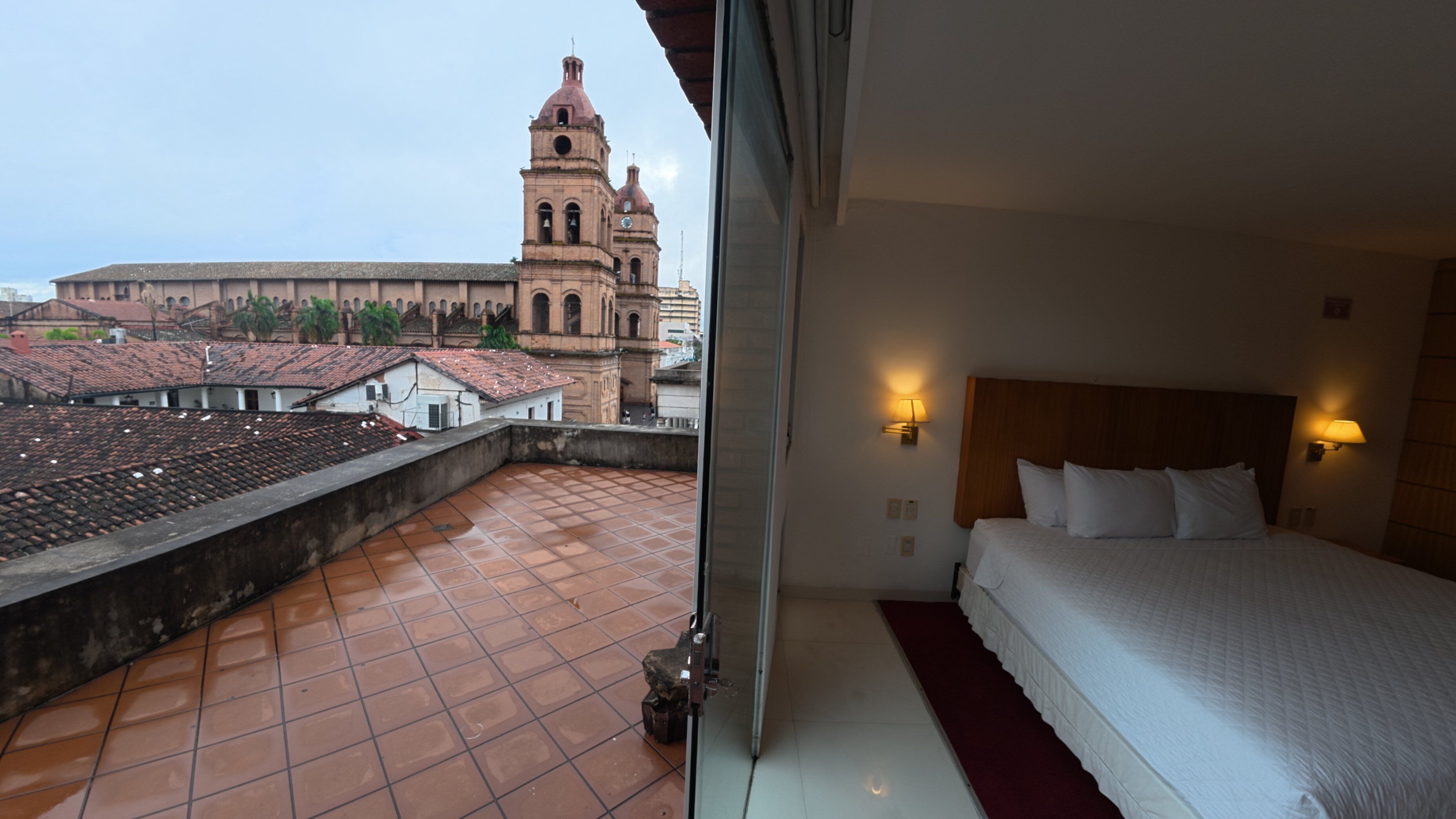 Quarto de hotel com uma cama, mesa de cabeceira e vista para um edifício histórico com um campanário.