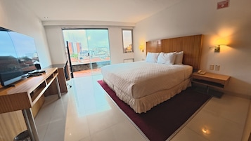Quarto de hotel com uma cama grande, uma escrivaninha com televisão, uma mesa de cabeceira e vista para a cidade pela janela.