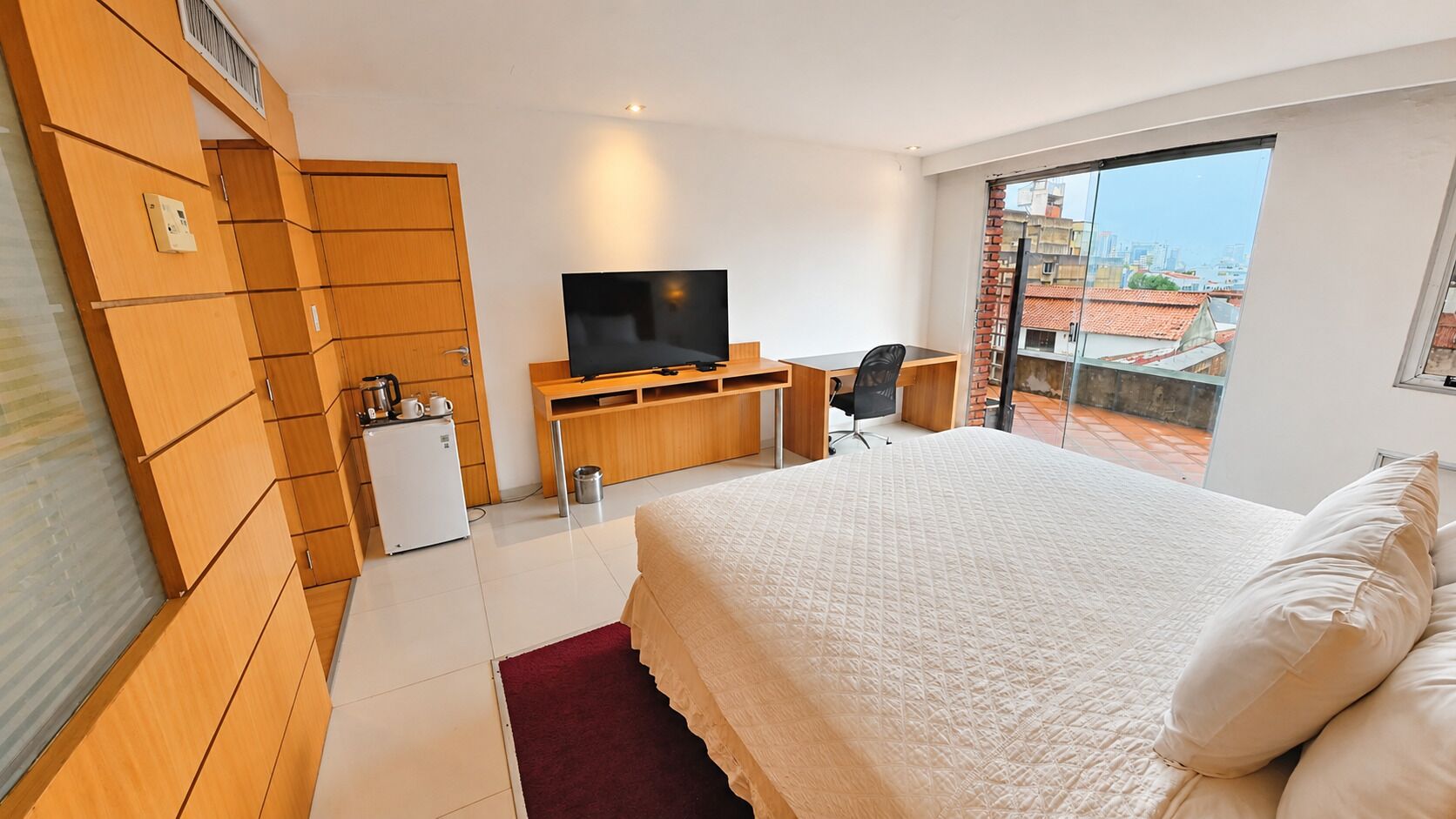 Quarto de hotel com cama, escrivaninha, televisão e vista da cidade por uma grande janela.