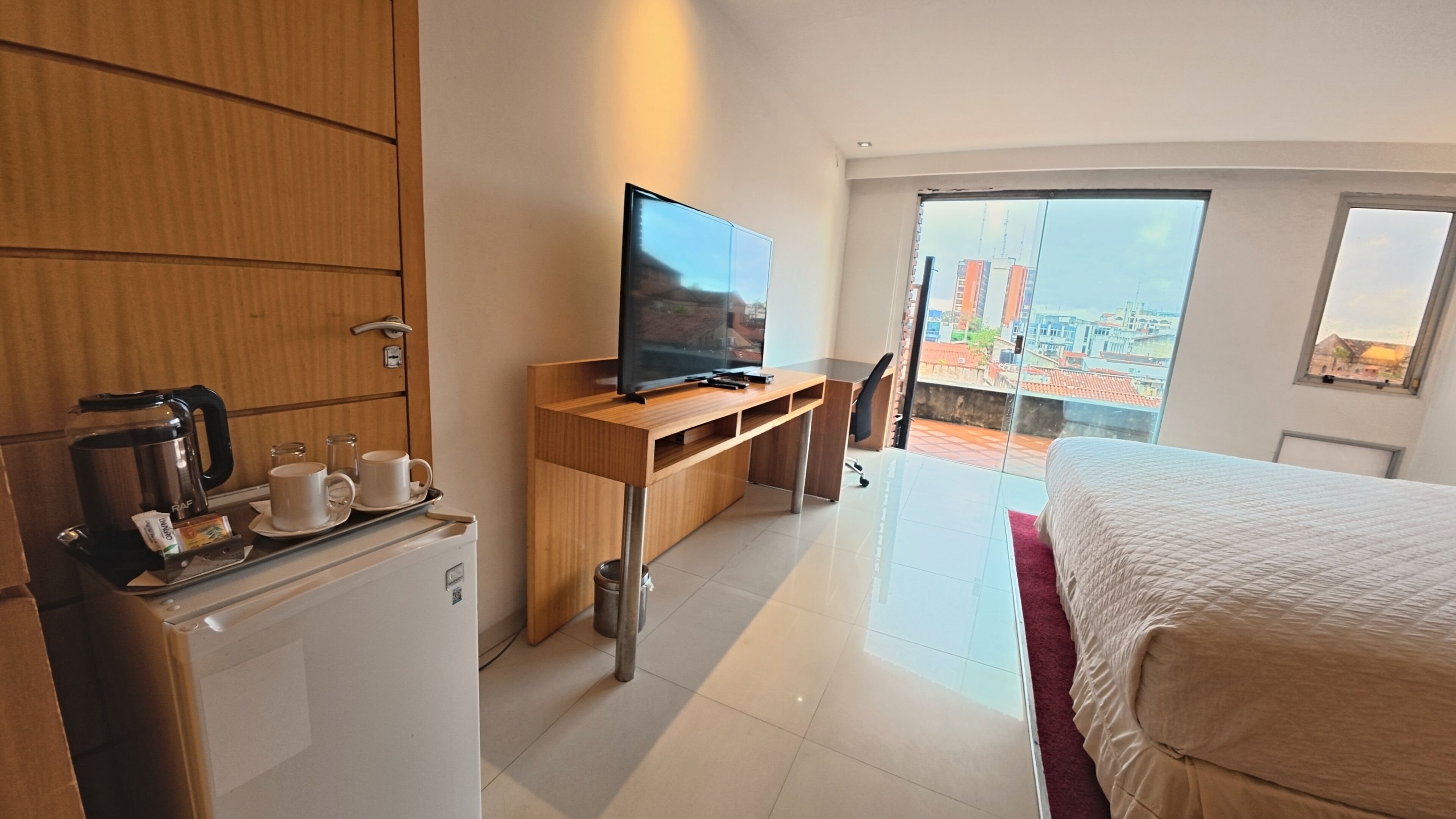 Quarto de hotel com cama, escrivaninha com televisão, cafeteira e vista da cidade através de grandes janelas.