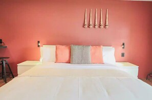 Uma cama com roupa de cama branca, um travesseiro estampado e uma parede vermelha com luminária.