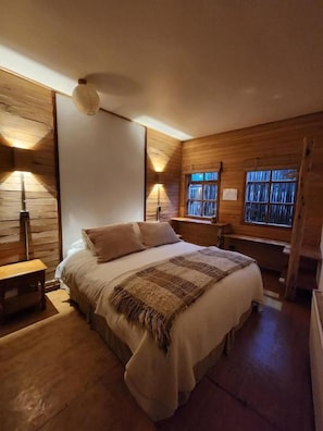 Quarto com uma cama grande, paredes de madeira e duas janelas.