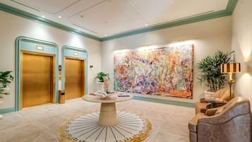 Um hall com dois elevadores, uma grande pintura abstrata, uma mesa redonda com livros e uma poltrona de couro.