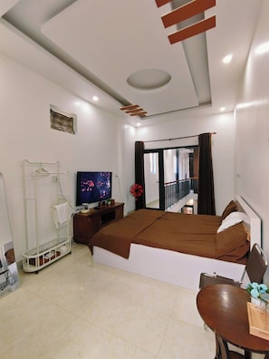 Quarto de hotel com cama, televisão, mesa de cabeceira, cadeira, pequena mesa com flores e janela com cortinas.