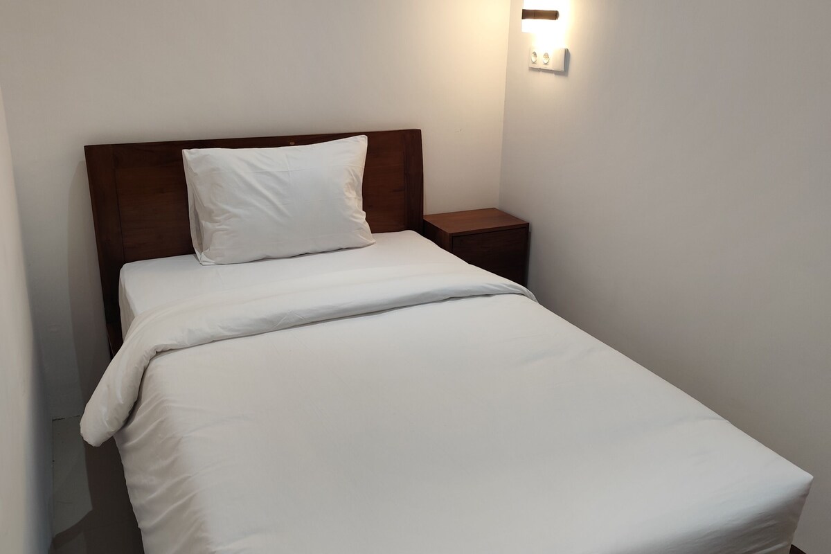 Uma cama de solteiro com cabeceira de madeira, mesa de cabeceira e luminária fixa na parede.