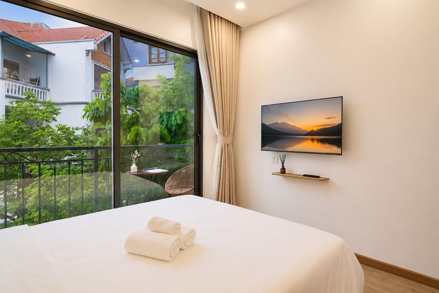 Quarto de hotel com cama, TV fixada na parede e varanda com vista para edifícios e vegetação.