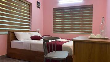 Um quarto com uma cama, uma escrivaninha e uma cadeira. Há duas janelas com persianas, um ar condicionado fixado na parede e uma pequena imagem na parede.