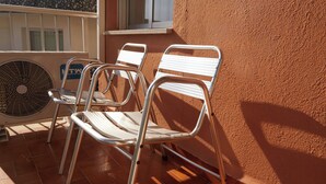 Duas cadeiras de metal com almofadas brancas, um ventilador grande e uma janela com vista para um edifício.