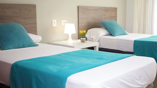 Quarto de hotel com duas camas, uma mesa de cabeceira com abajur e uma pequena mesa com um vaso de flores.