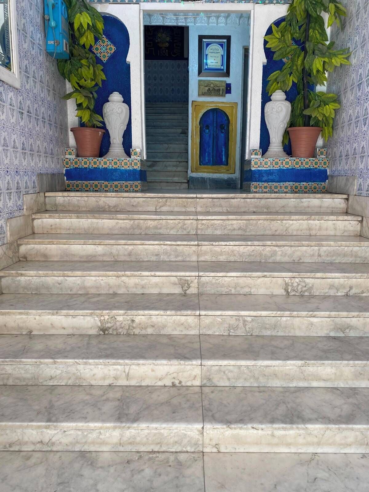 Um conjunto de degraus de mármore que levam a uma porta azul com uma placa, ladeada por plantas em vasos e azulejos decorativos.