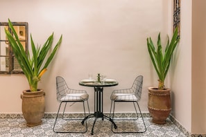 Um canto com duas cadeiras de metal, uma mesa redonda pequena e duas plantas em vasos com folhas verdes longas.