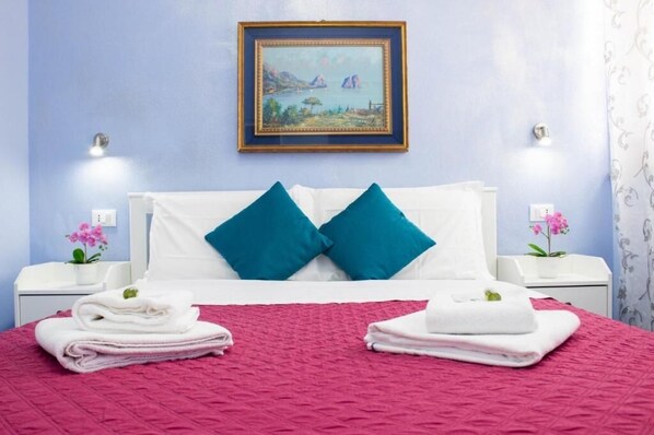 Quarto de hotel com cama, almofadas, um quadro na parede e duas mesas de cabeceira com abajures e flores.