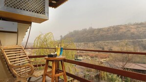 A balcony with a wooden chair, a small table with a drink, and a view of a hilly landscape.