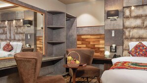 A hotel room with a large mirror, a wooden headboard, a brown armchair, a bedside table with a basket of fruit, and a bed with a colorful blanket.