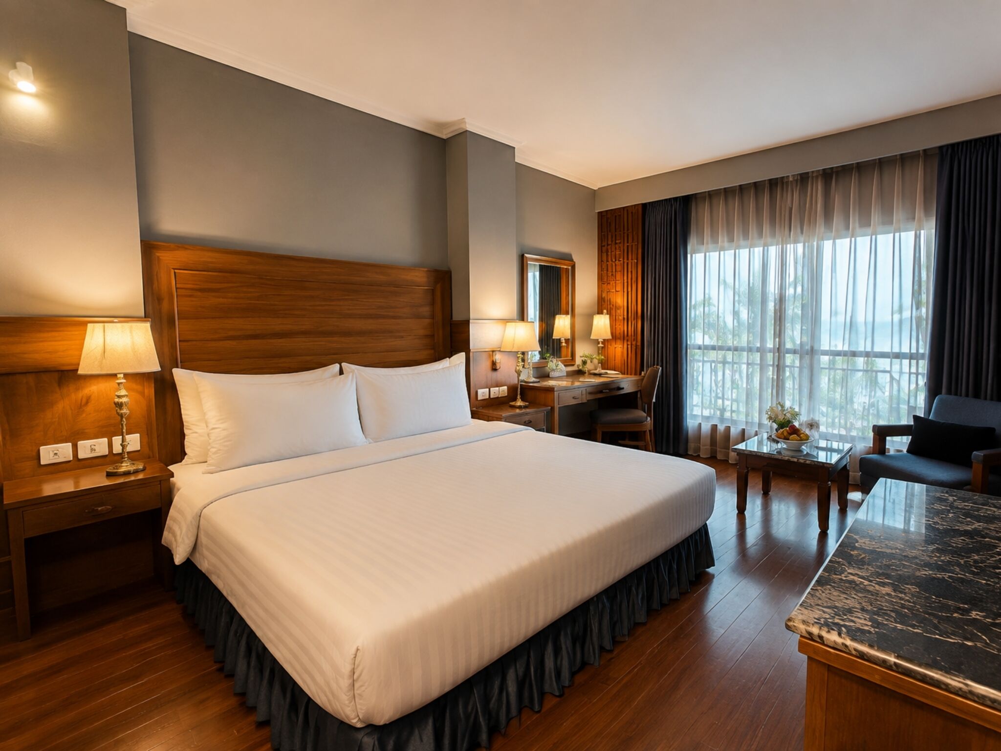 Quarto de hotel com uma cama grande, luminárias de cabeceira, uma escrivaninha com cadeira, um espelho e vista para o exterior pela janela.