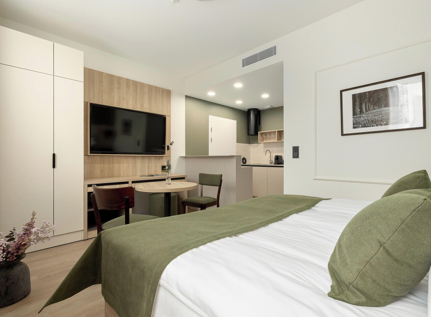 Quarto de hotel moderno com cama, secretária, cadeira, televisão e cozinha compacta.
