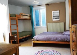 Quarto com beliches, uma cama de solteiro, cômoda de madeira, radiador e janela com cortinas.
