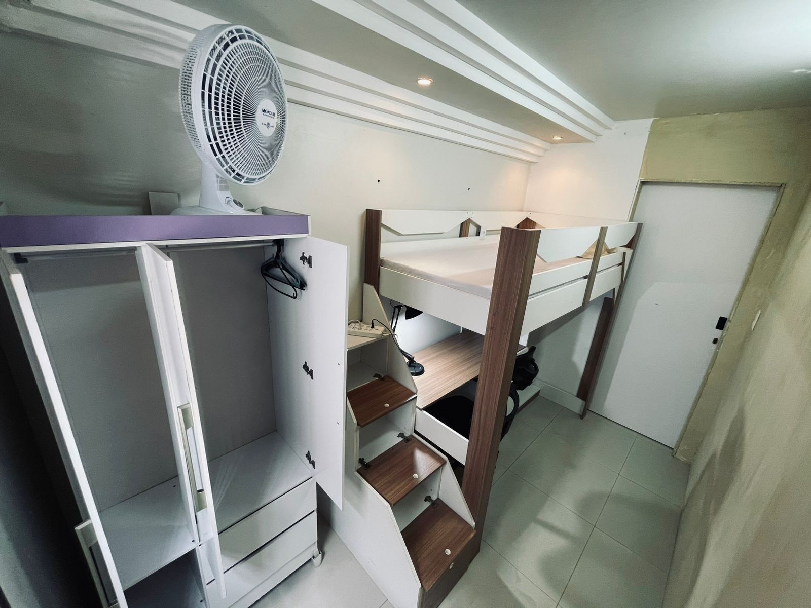 Quarto compacto com beliches, guarda-roupa, escrivaninha e ventoinha no teto.
