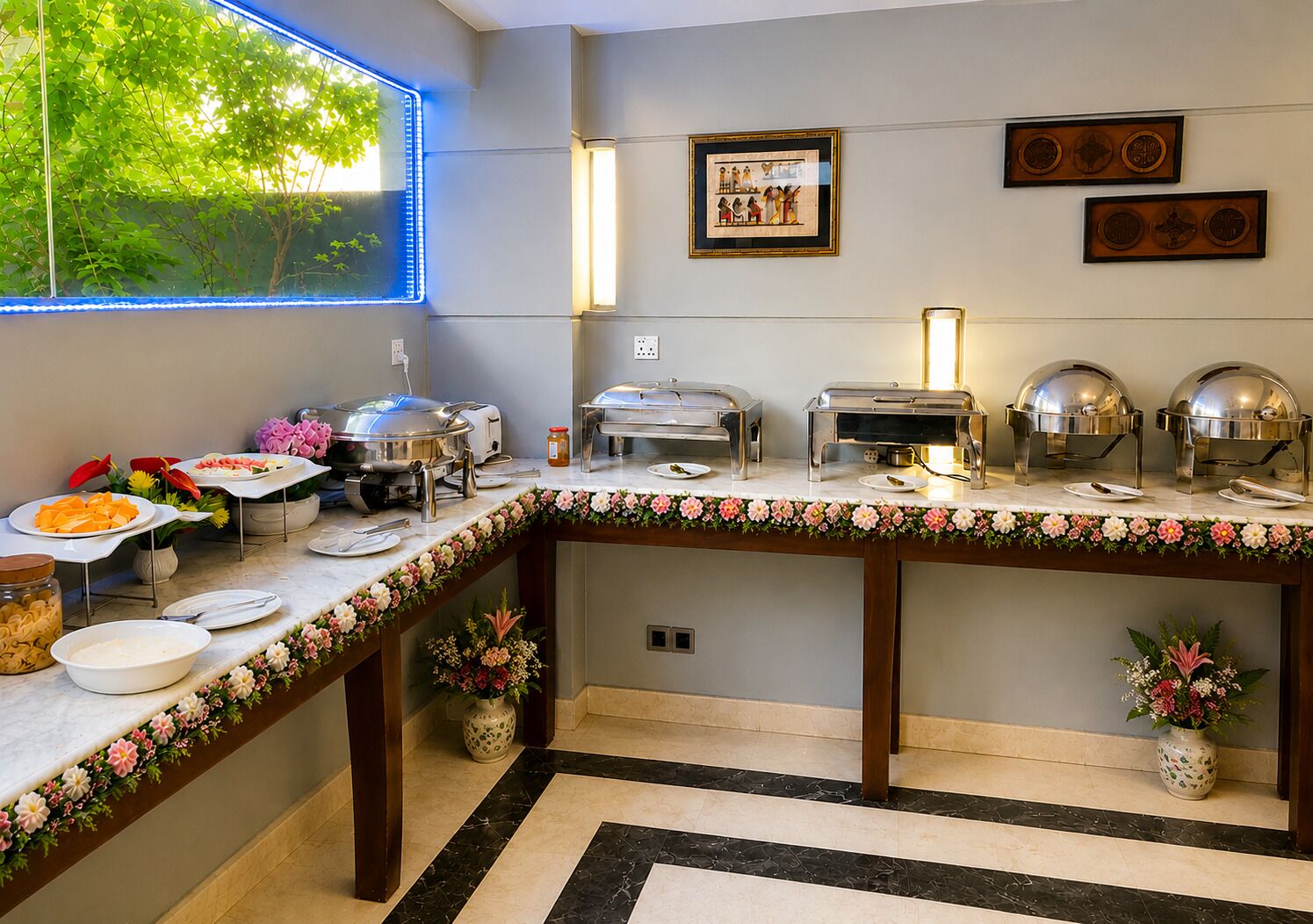 Disposição de buffet com diversos alimentos e estações de serviço, uma moldura na parede e uma janela com vista para o verde.