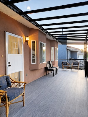 Uma área externa coberta com um deck de madeira, duas cadeiras de vime, uma porta branca com janelas protegidas por grades e um acessório de luz fixado na parede.