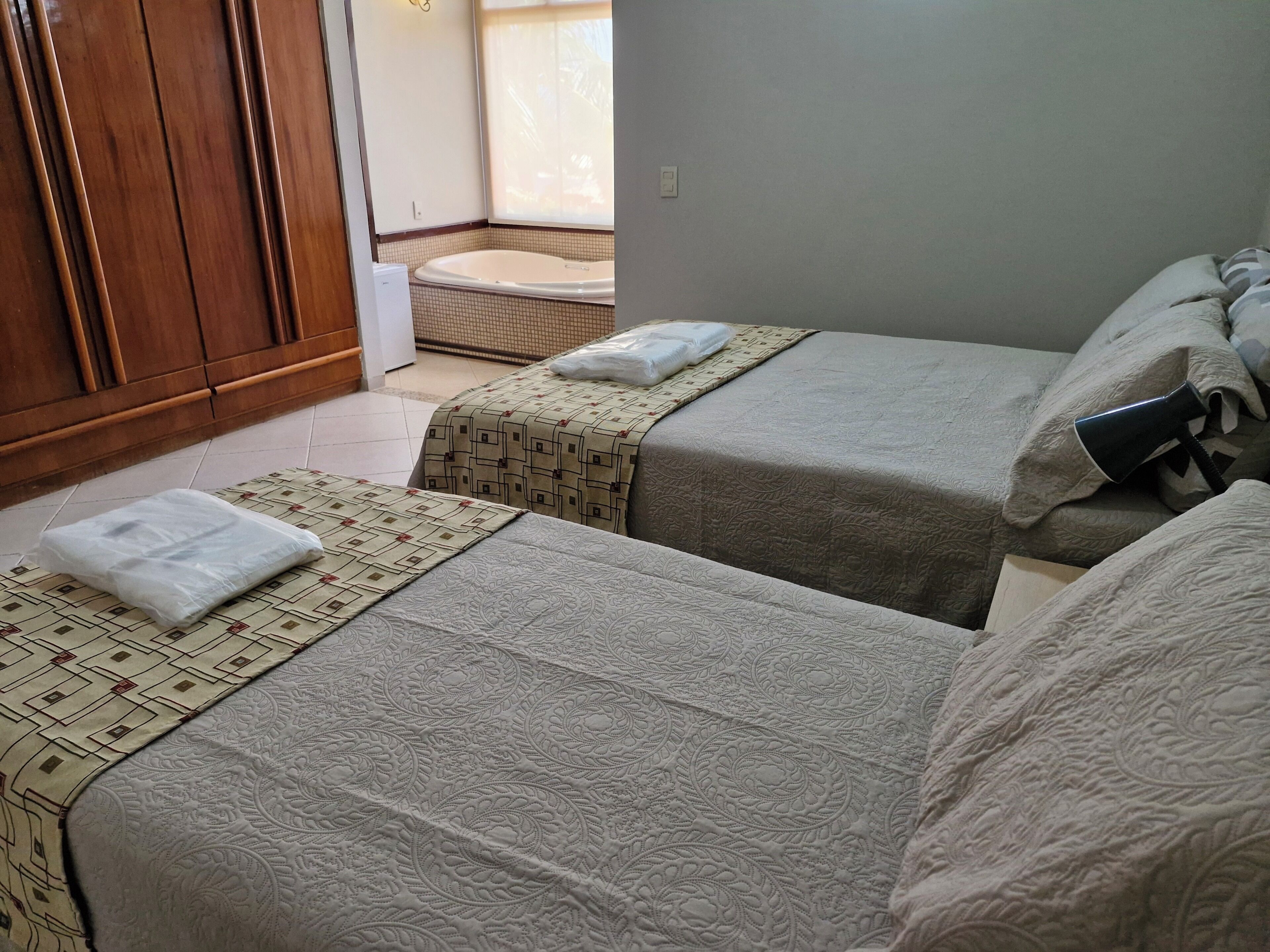 Quarto de hotel com duas camas, um guarda-roupa de madeira, banheiro com banheira e janela com cortinas.