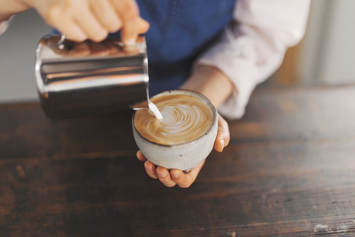 Pessoa segurando uma xícara de café com arte latte, usando um bule de metal para servir.