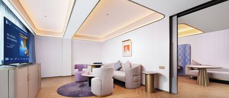 Quarto de hotel moderno com televisão de tela plana, sofá, mesa de centro e pequena mesa lateral.