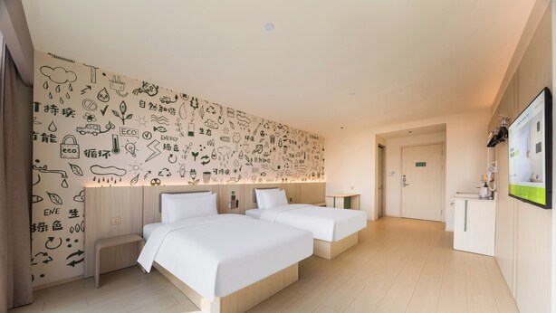 Quarto de hotel com duas camas, parede decorada com desenhos de temática ecológica, uma pequena mesa de cabeceira e uma área de cozinha compacta.