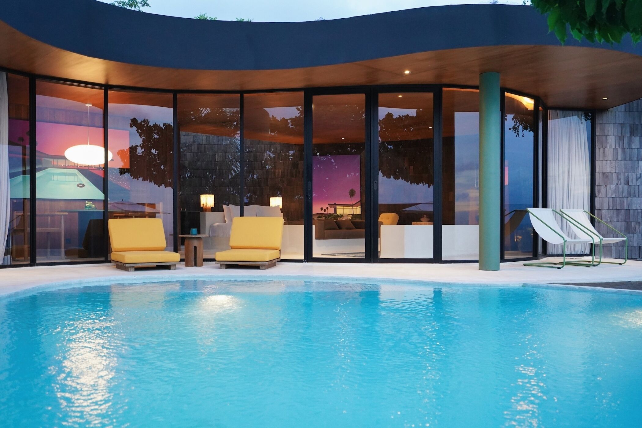 Piscina interior moderna com paredes de vidro transparente, cadeiras de descanso amarelas e vista para o pôr do sol.