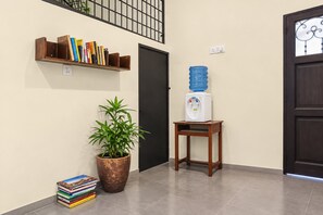 Corredor com estante, bebedouro, planta em vaso e pilha de livros.