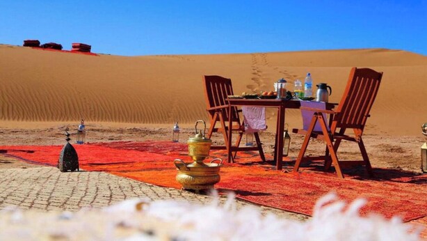 Montagem de refeição no deserto com mesa, cadeiras, tapete vermelho e uma duna ao fundo.